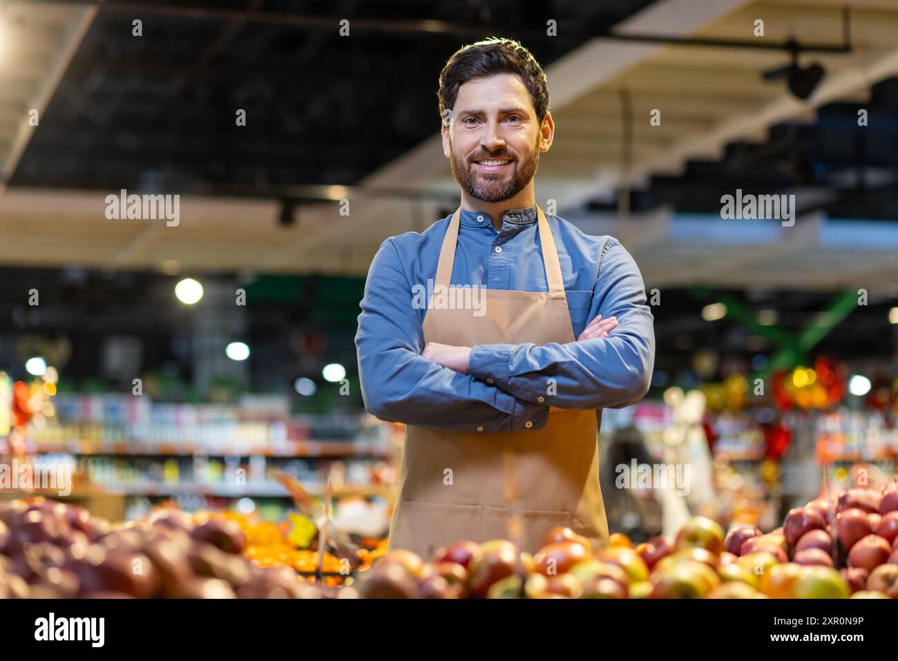 Confident male grocery store worker wearing apron standing with arms ...