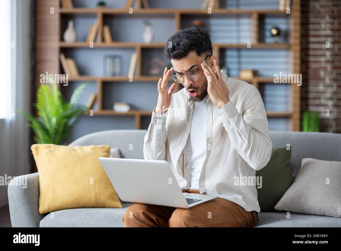Shocked man sitting on couch with laptop on lap, hands on head ...