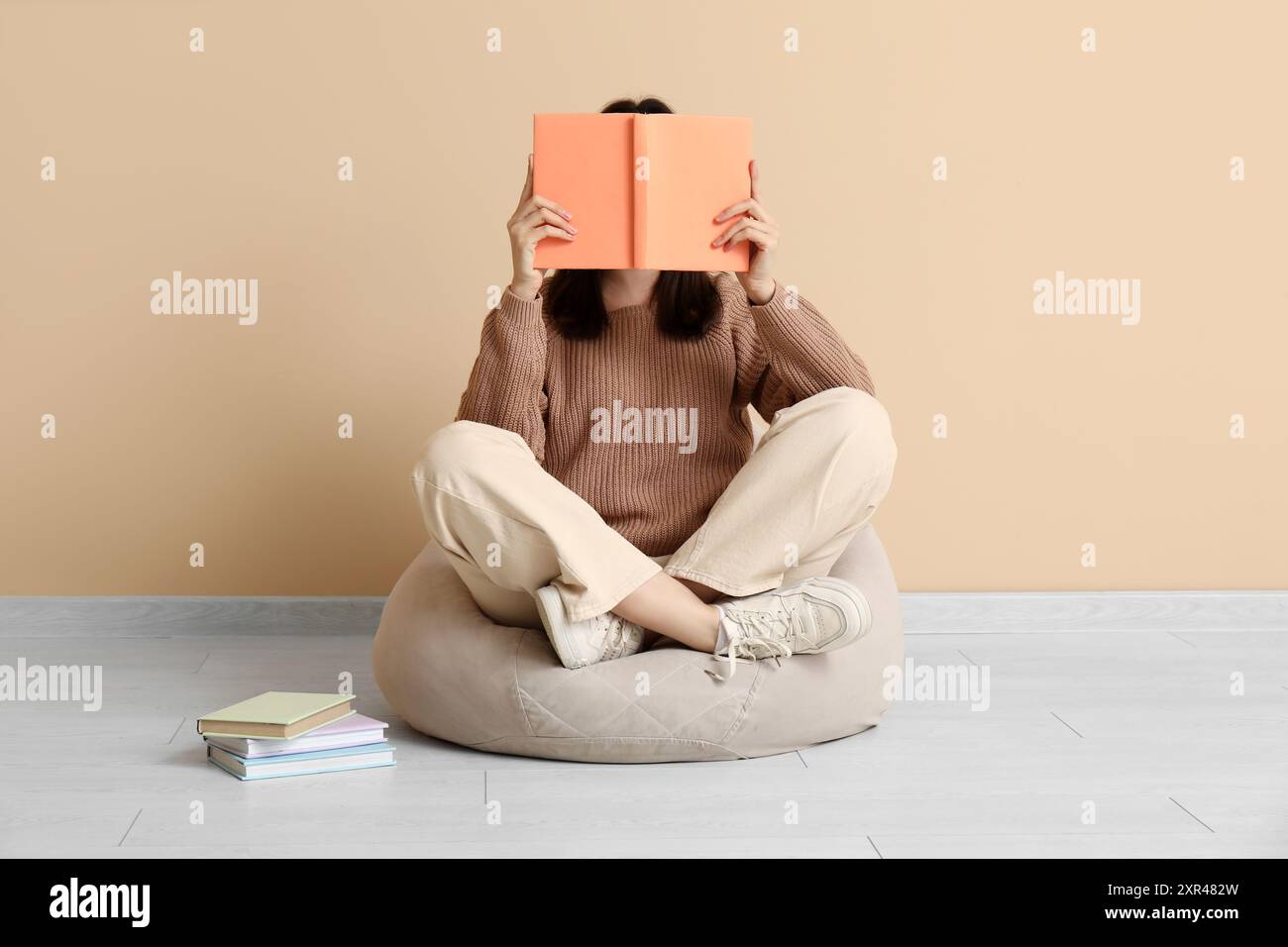 Young woman sitting on pouf and reading book near beige wall Stock ...
