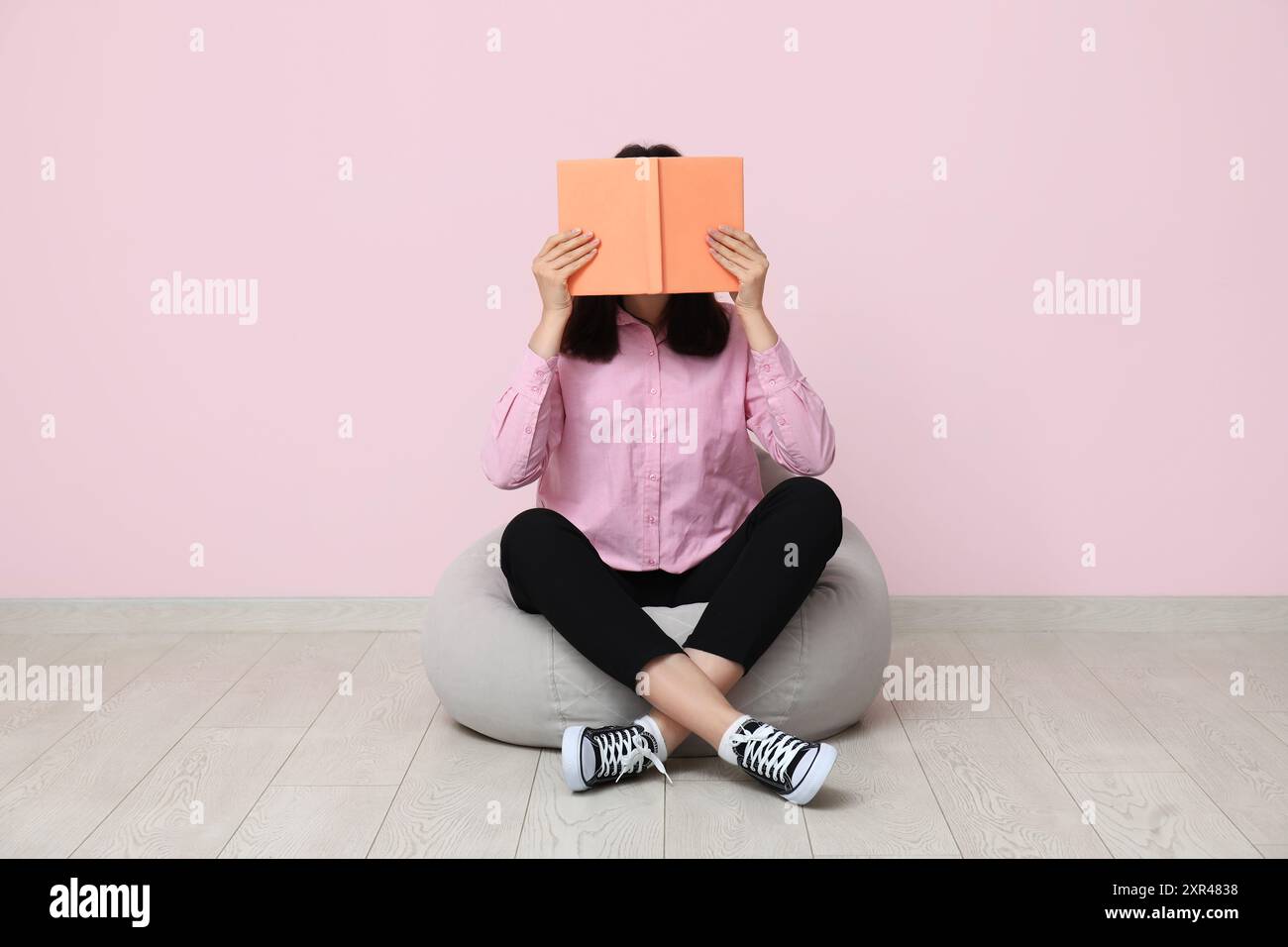 Young woman sitting on pouf and reading book near pink wall Stock Photo ...
