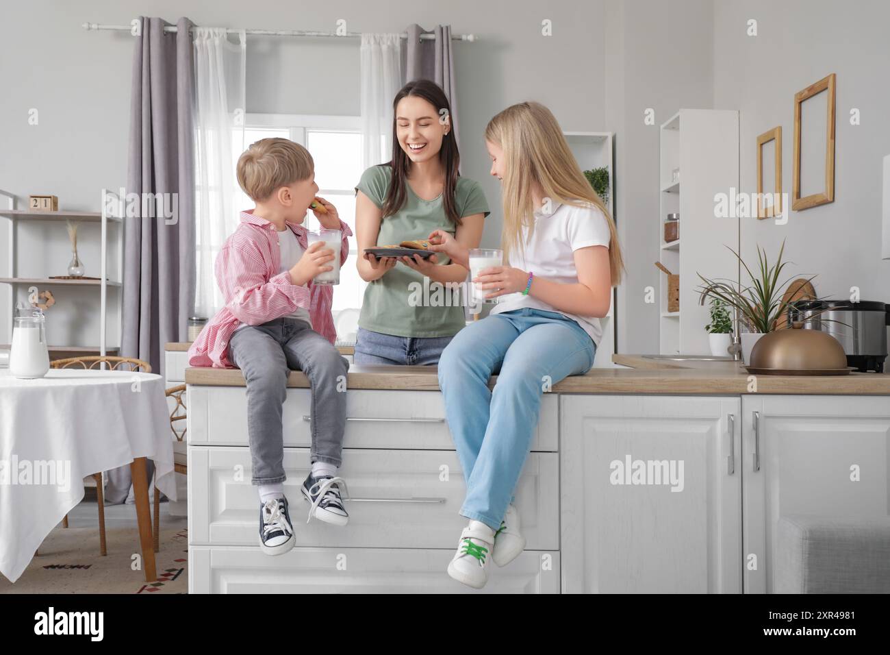 Mom giving cookies to her children sitting on counter and drinking milk ...