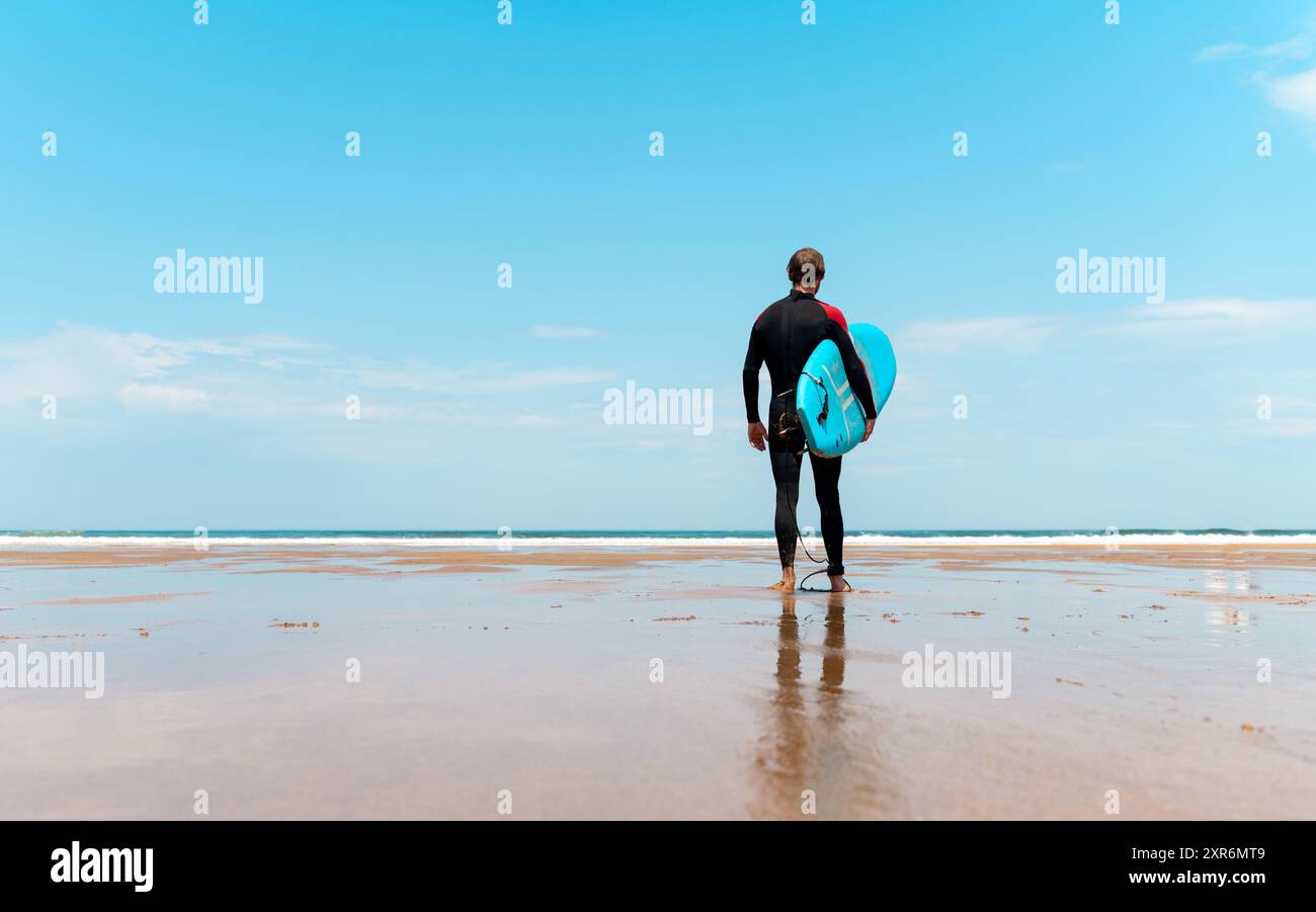 Back view of a young surfer man in wetsuit and surfboard on the beach ...