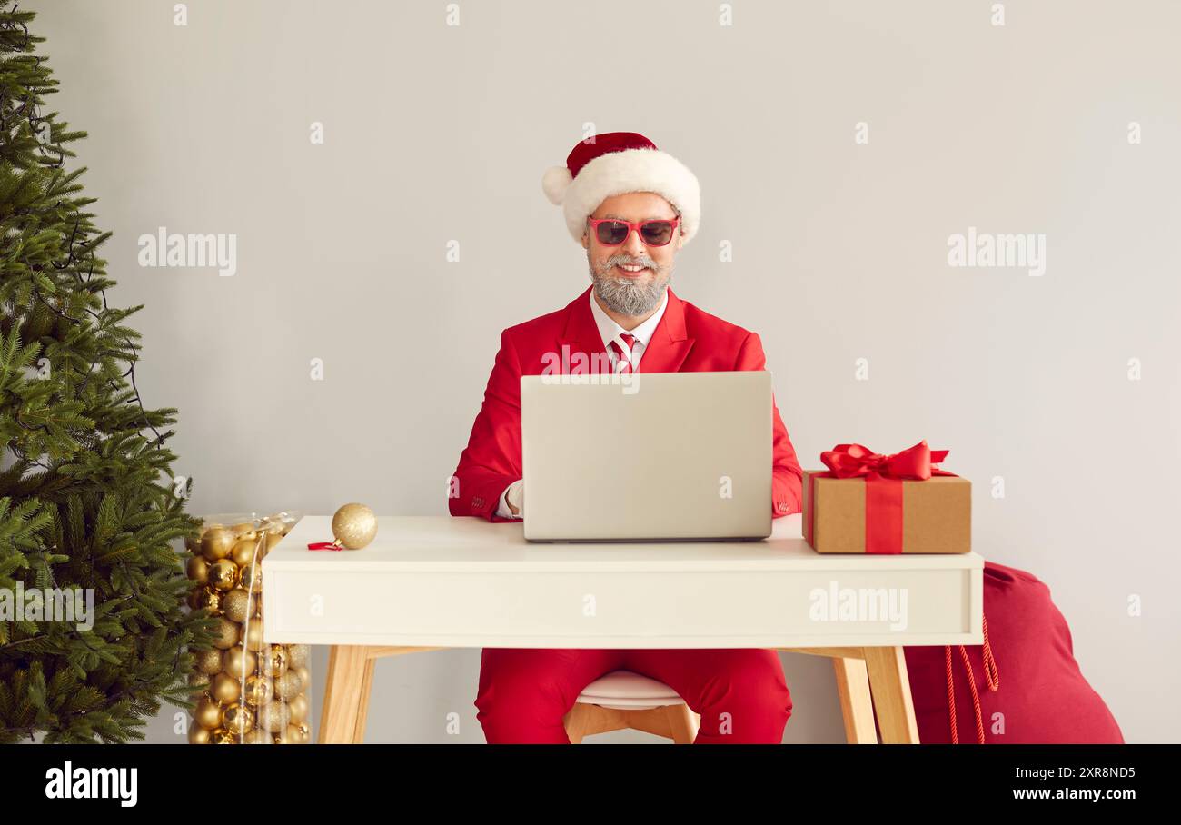 Funny modern Santa working on laptop while sitting at office desk next ...