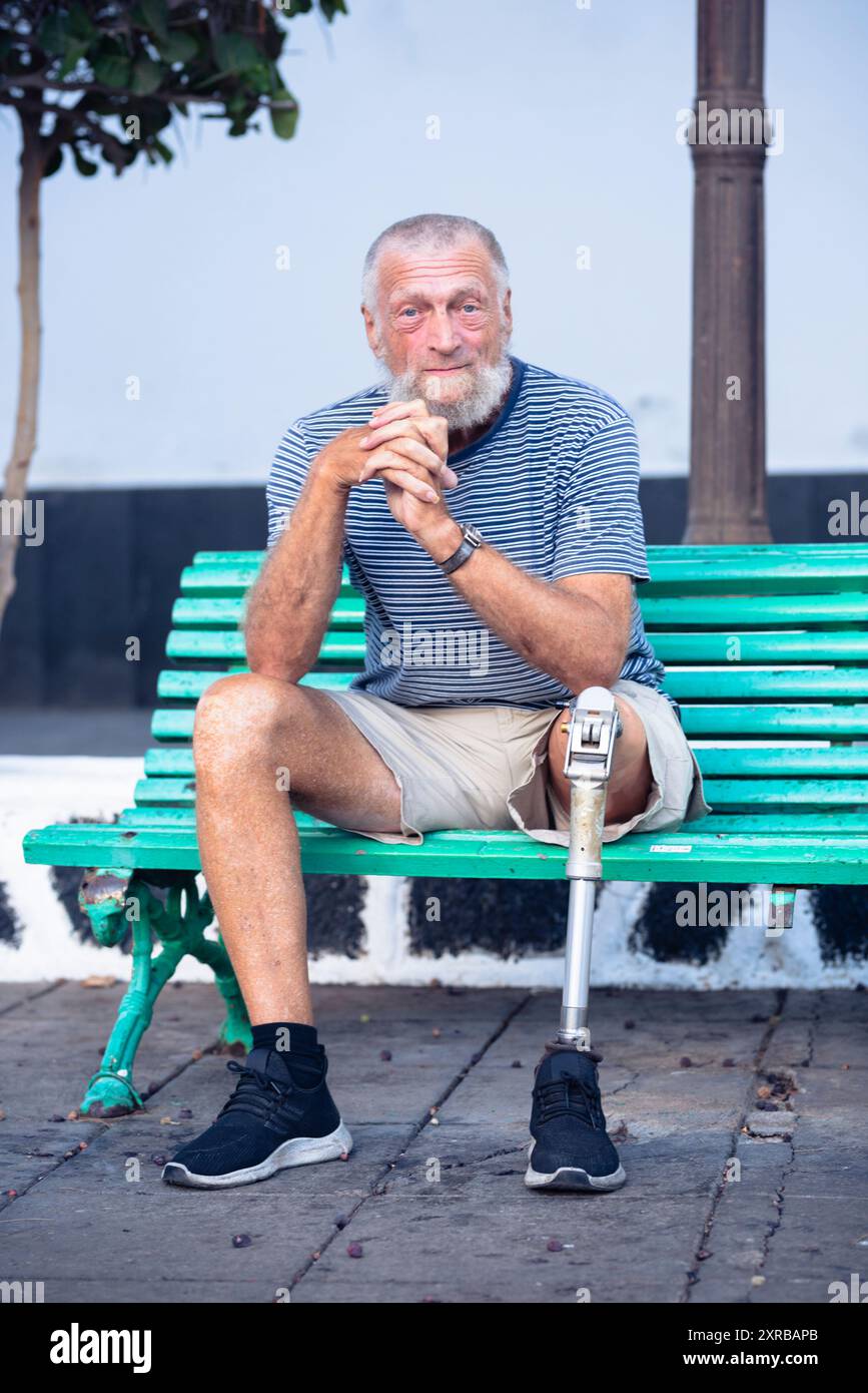 Elderly man with prosthetic leg left rests sitting on a park bench ...