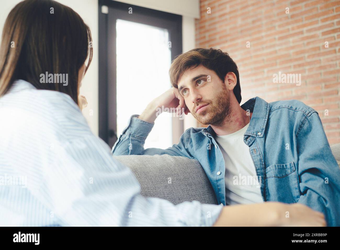 A young 25-year-old couple sits on their cozy sofa in a living room, engaged in a quiet ...