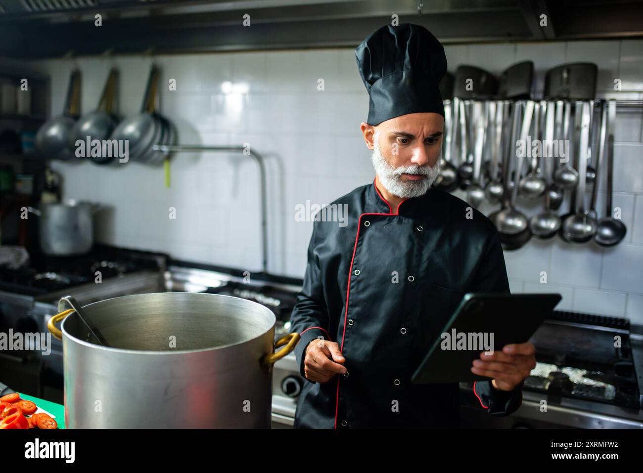 Mature chef in traditional uniform focusing on a digital tablet while ...