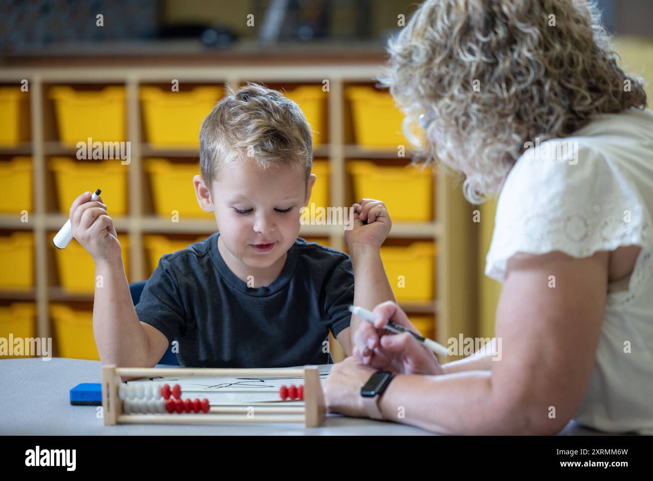 A young boy is sitting at a table with a teacher, who is helping him ...