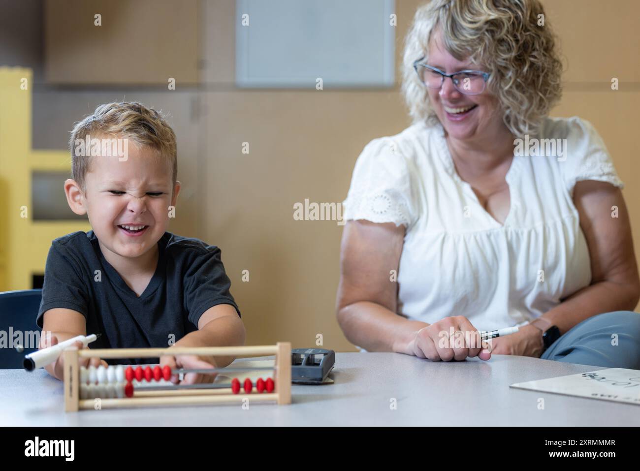 A young boy is sitting at a table with an abacus and smiling with his teacher laughing with him ...