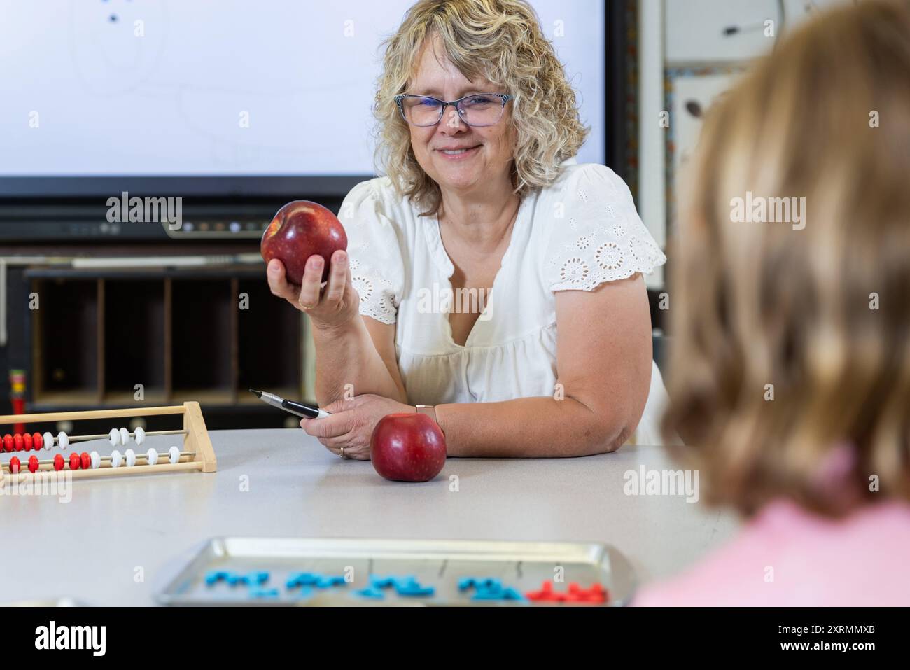 A teacher is holding an apple and smiling at a child. The teacher is ...