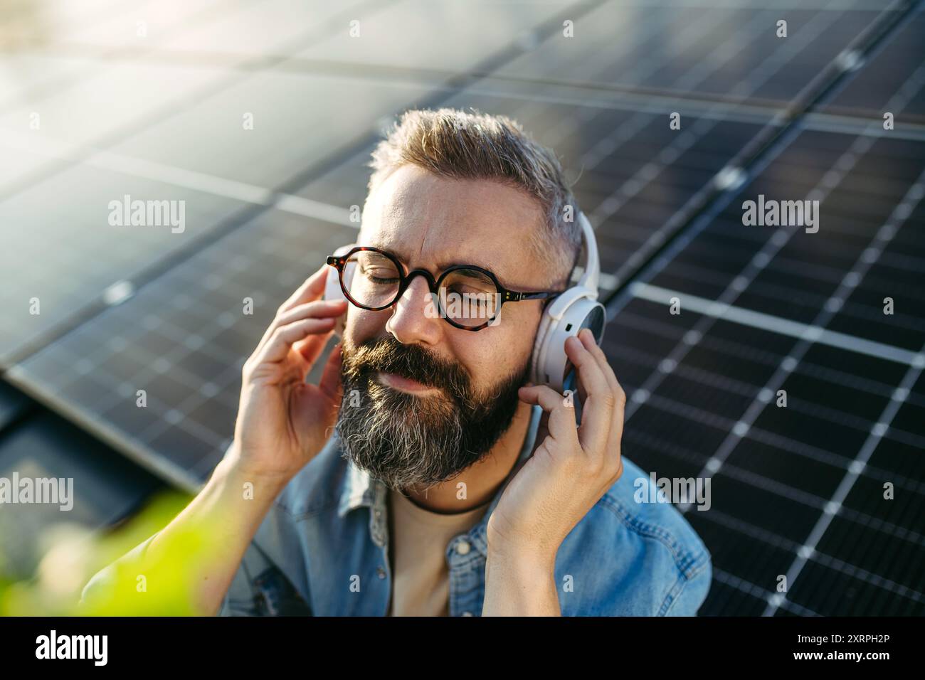 Man sitting on roof full of solar panels, headphones on head and closed ...