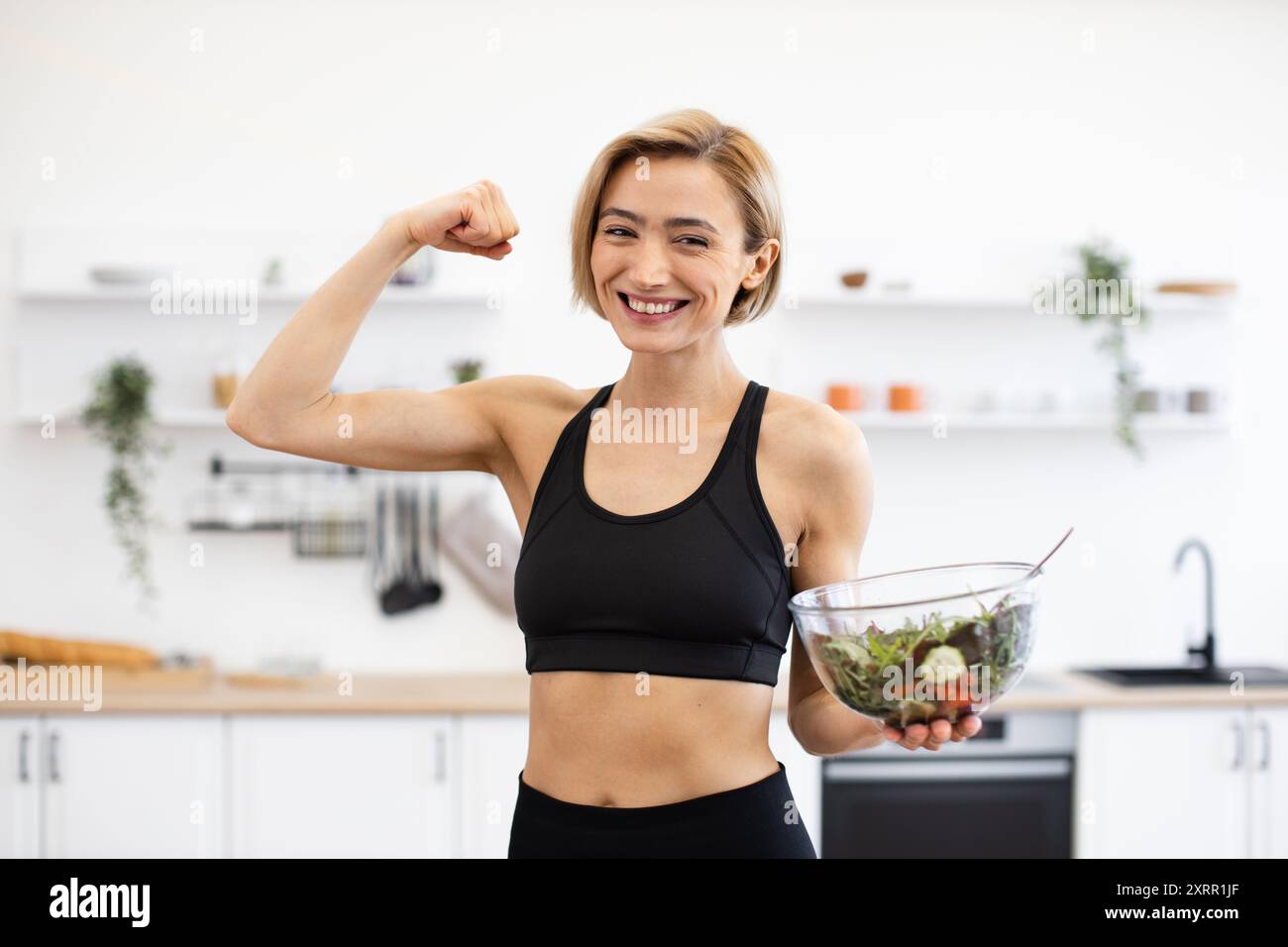 Healthy woman flexing muscles and holding salad in kitchen Stock Photo ...