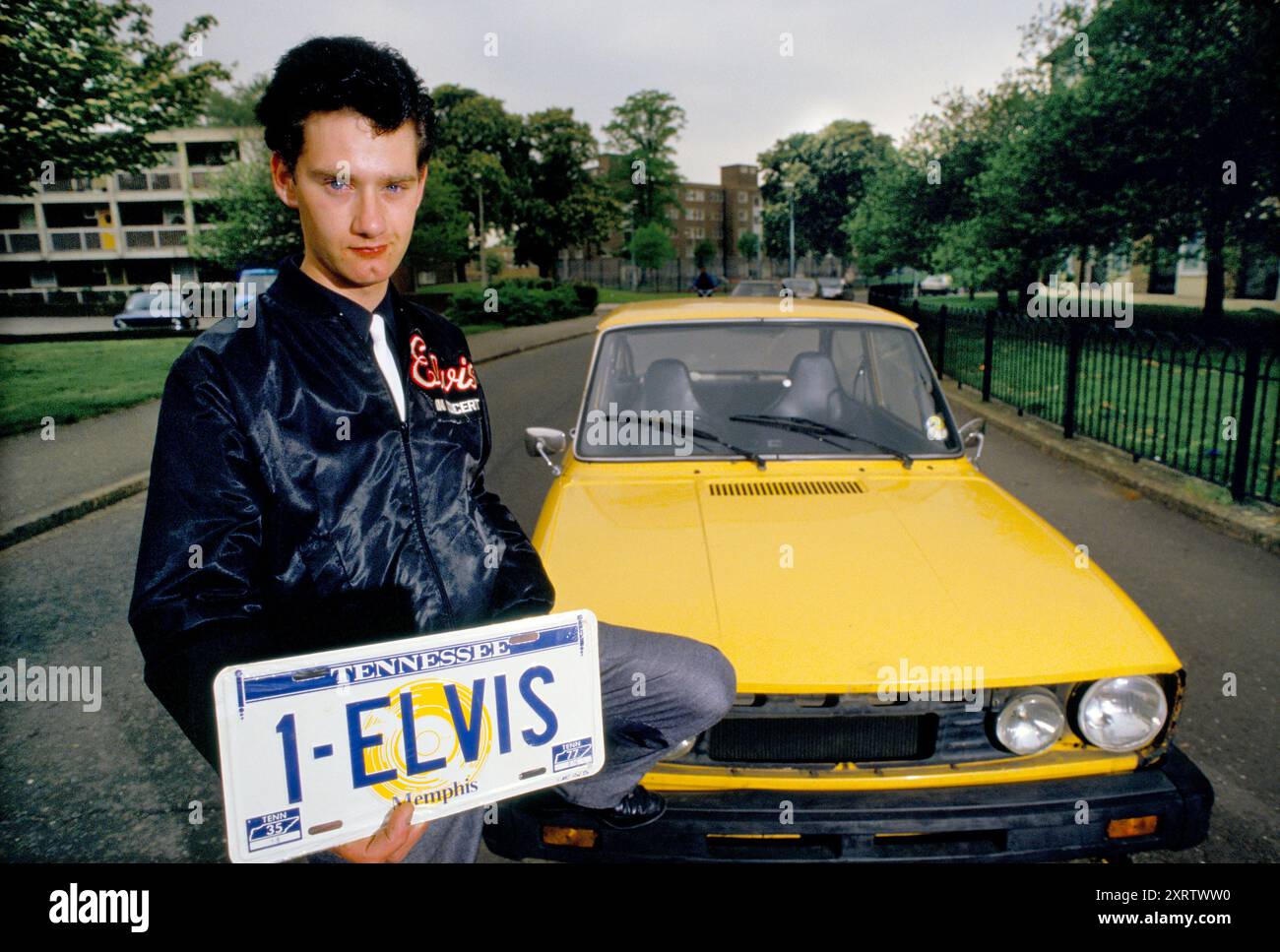 Elvis Presley fan, Chris standing in the street beside his bright yellow car holding his ...