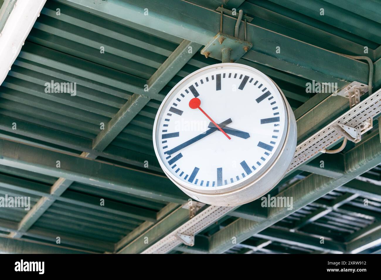A large clock hanging from a metal ceiling under a train station canopy ...