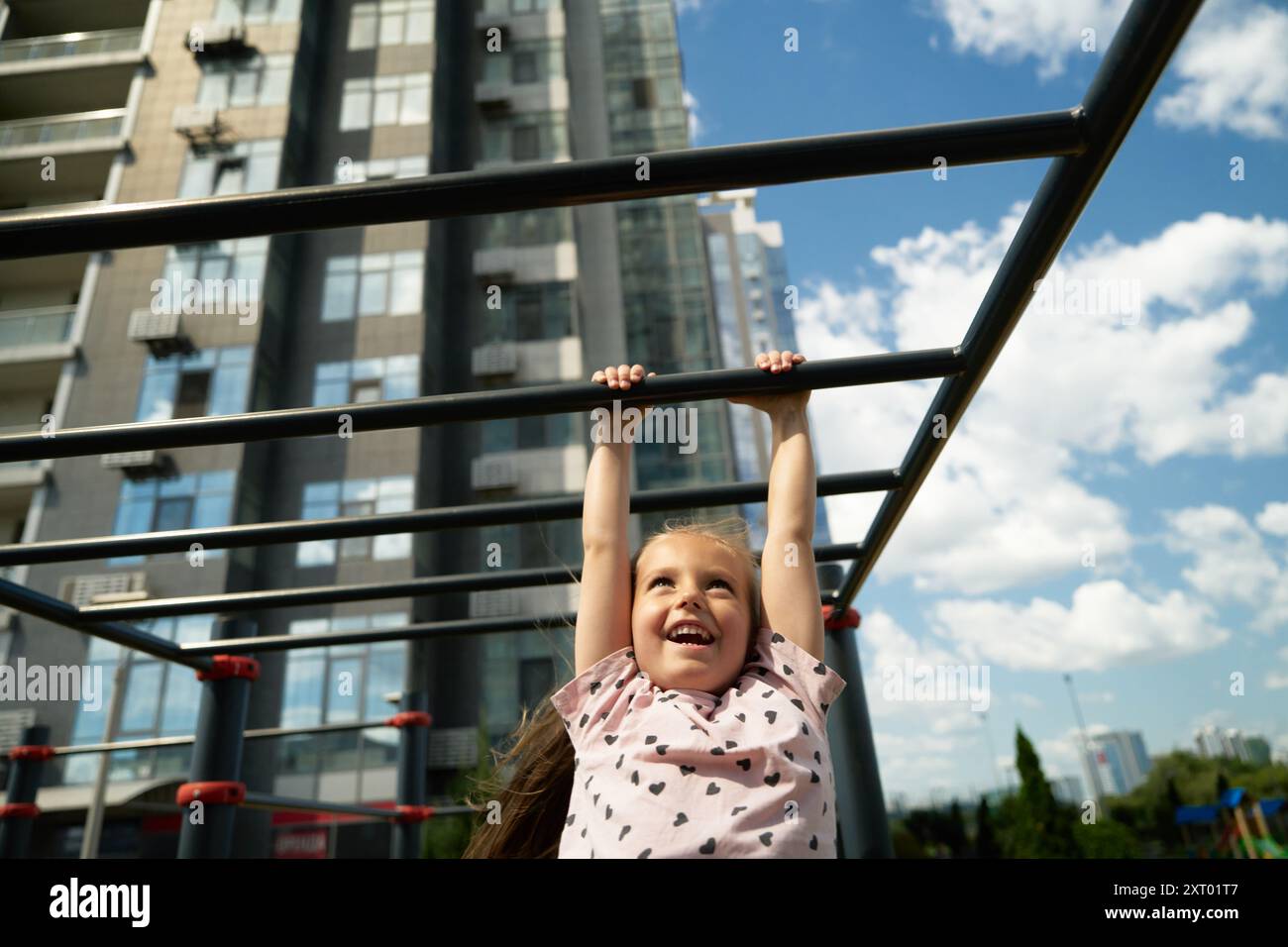 Young girl smiling while playing on monkey bars outside a tall building ...