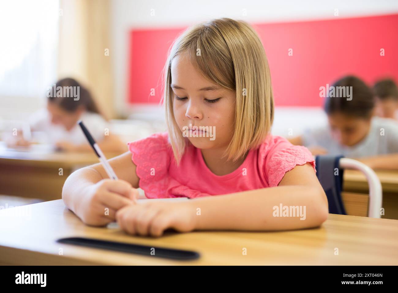 Interested blonde tween girl listening to schoolteacher and writing in ...