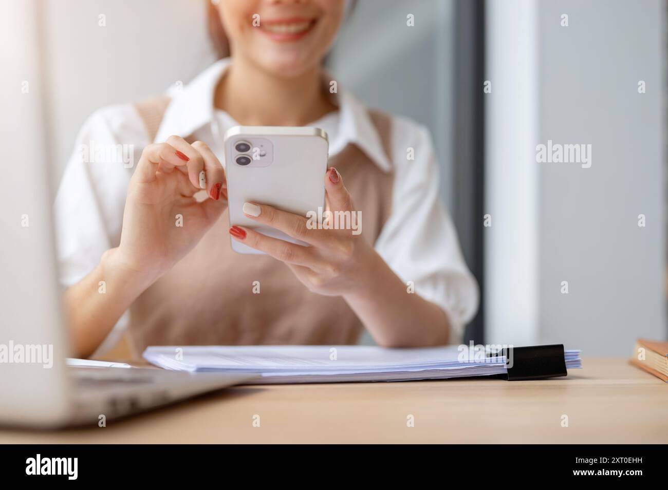 A cropped image of a beautiful, smiling Asian businesswoman using her smartphone at her desk in ...