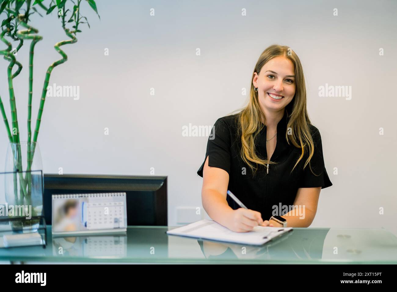 A smiling receptionist, dressed in a black uniform, sits behind a sleek ...