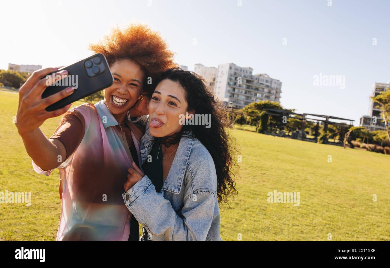 Two teenagers taking a playful selfie outdoors, one making a silly face ...