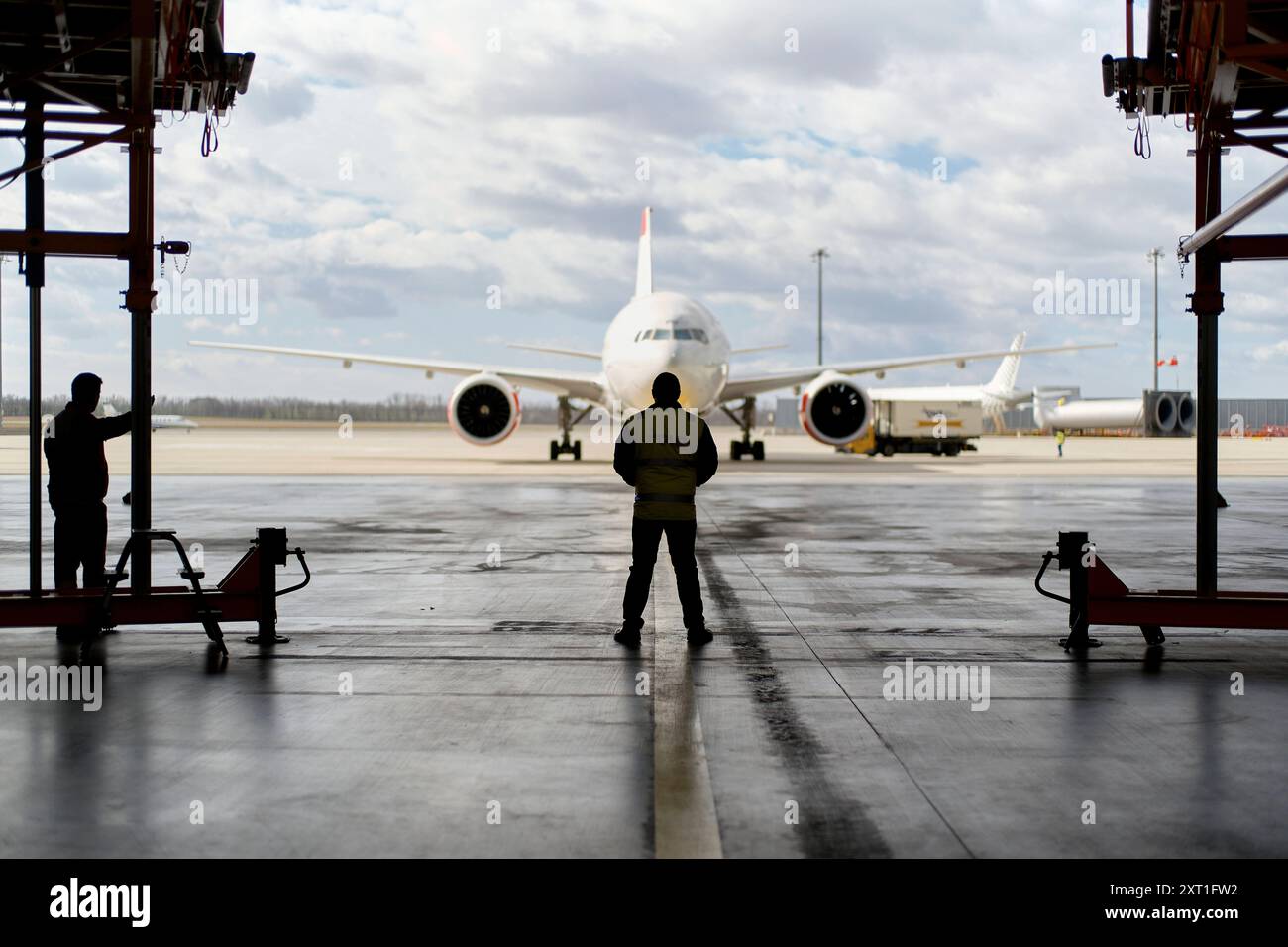 Ground crew member stands in an airplane hangar facing a parked commercial aircraft with another ...