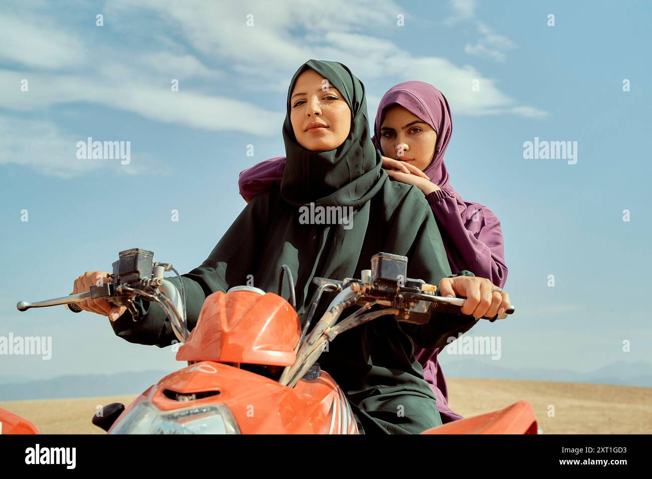 Two women in hijabs riding a red quad bike on sandy terrain under a clear sky. bola02555 ...