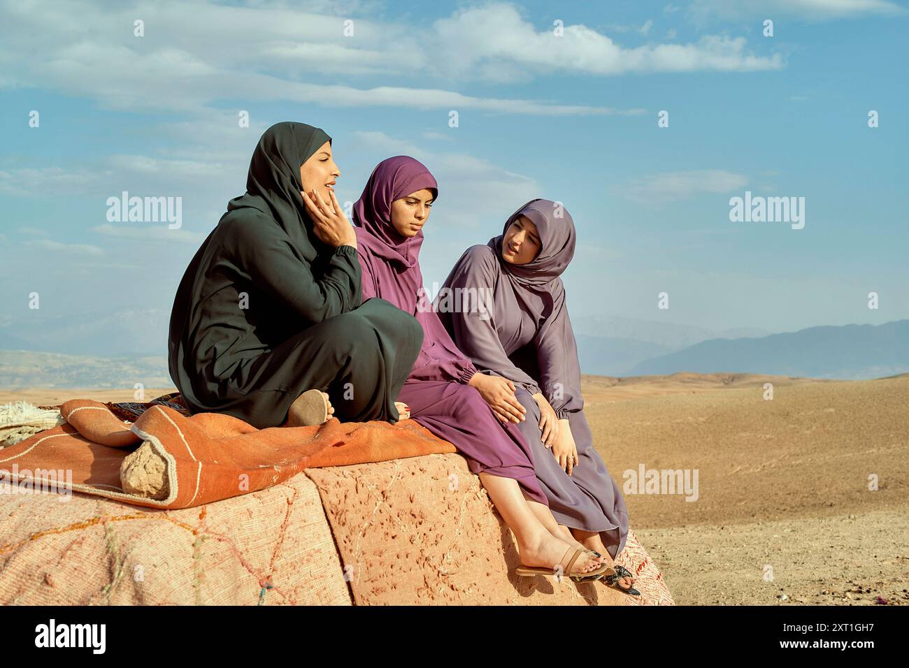 Three women in traditional hijab sitting together on a woven rug ...