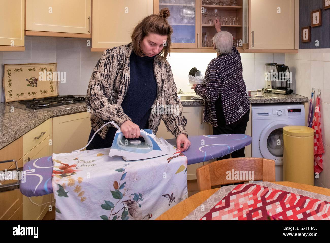 Ouderdom A woman irons clothes on an ironing board while another woman ...