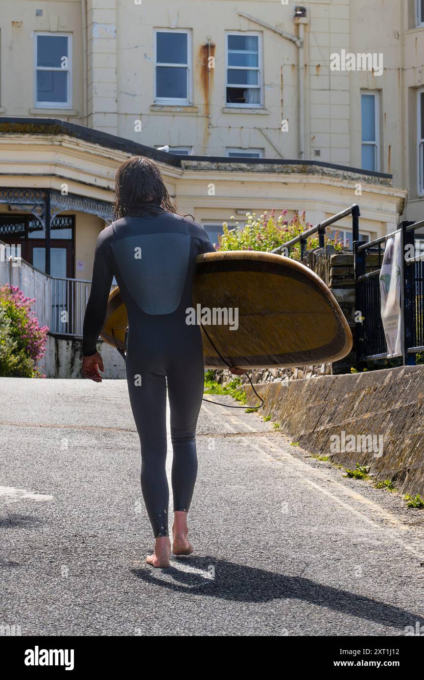 Rear view of a male surfer carrying his surfboard walking up the road ...