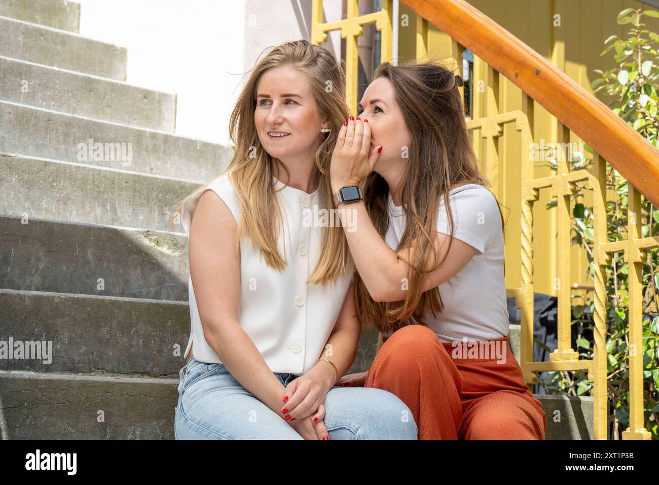 Two women sitting on outdoor steps, one whispering into the other s ear ...