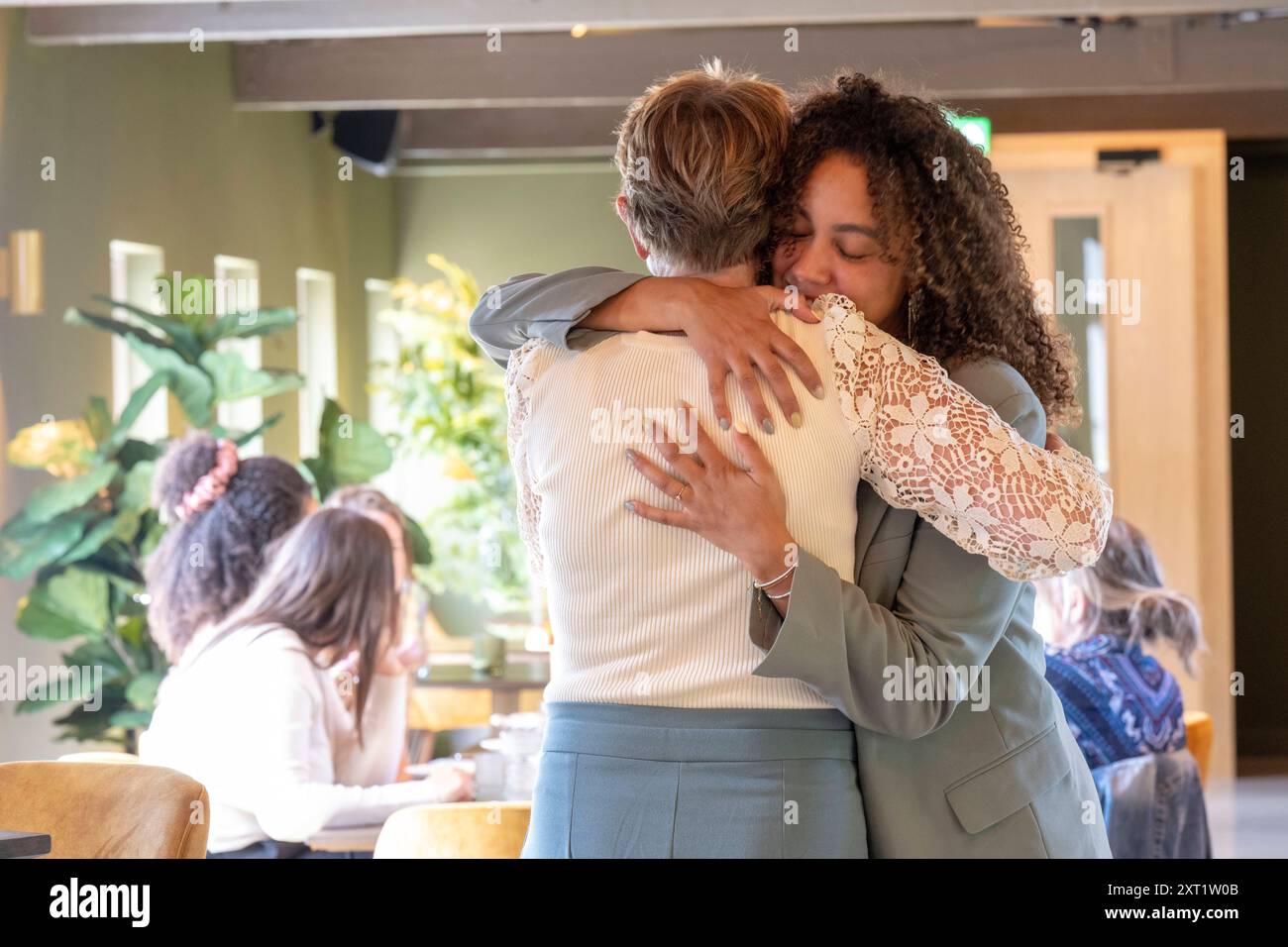 Two women embrace warmly in a cosy cafe setting as other patrons dine ...