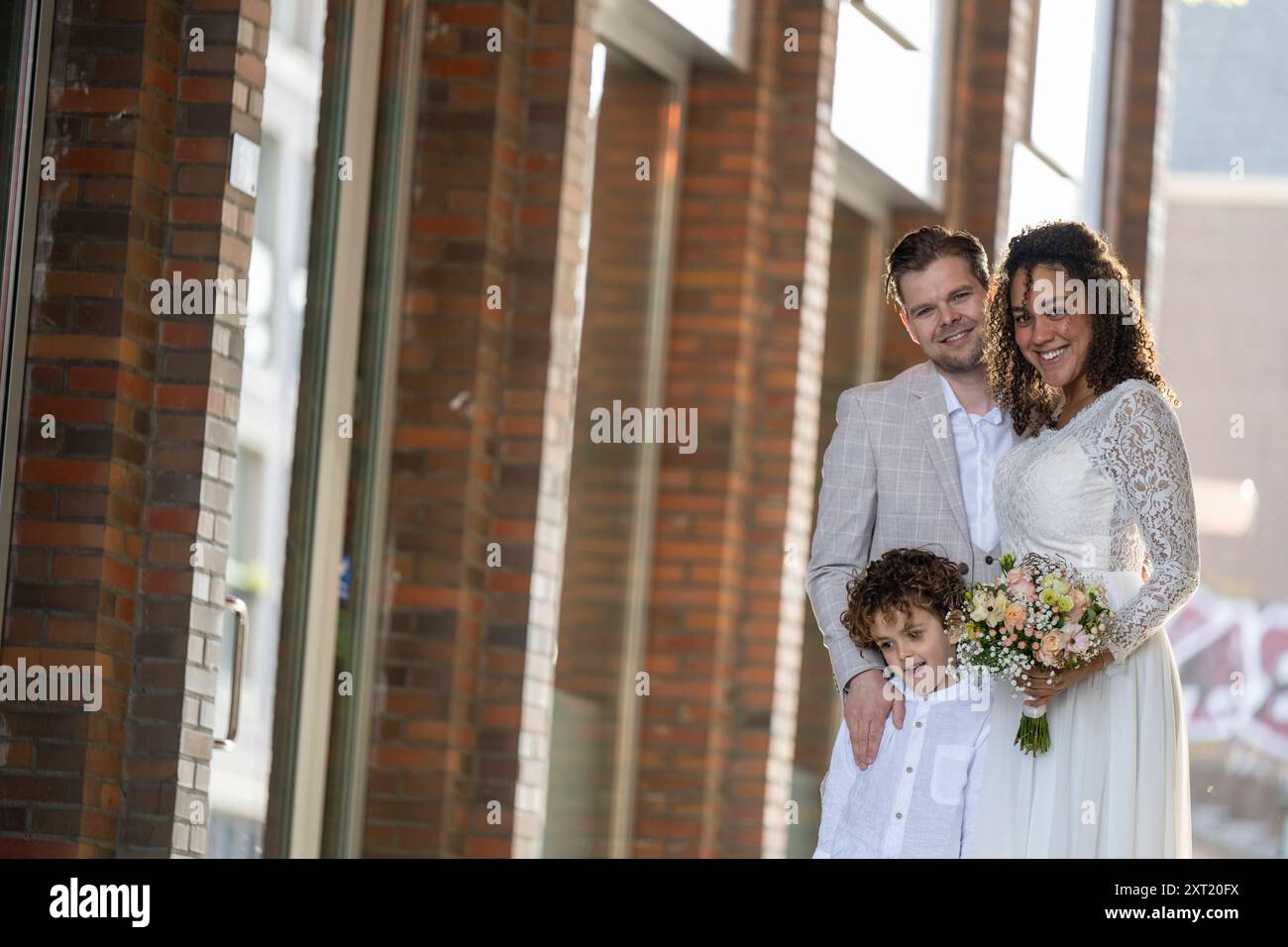 A smiling bride and groom pose with a young child, all dressed in ...