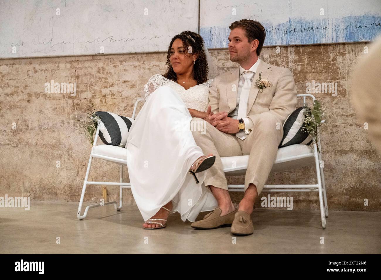A bride and groom sit closely together on a white bench, smiling ...