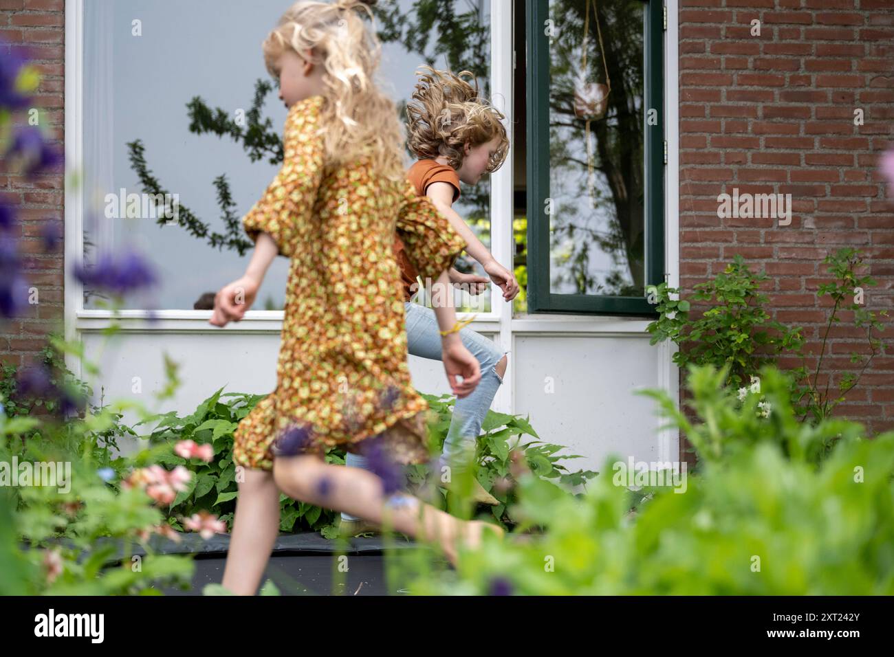 Two children playfully running in a garden with a red brick house in ...