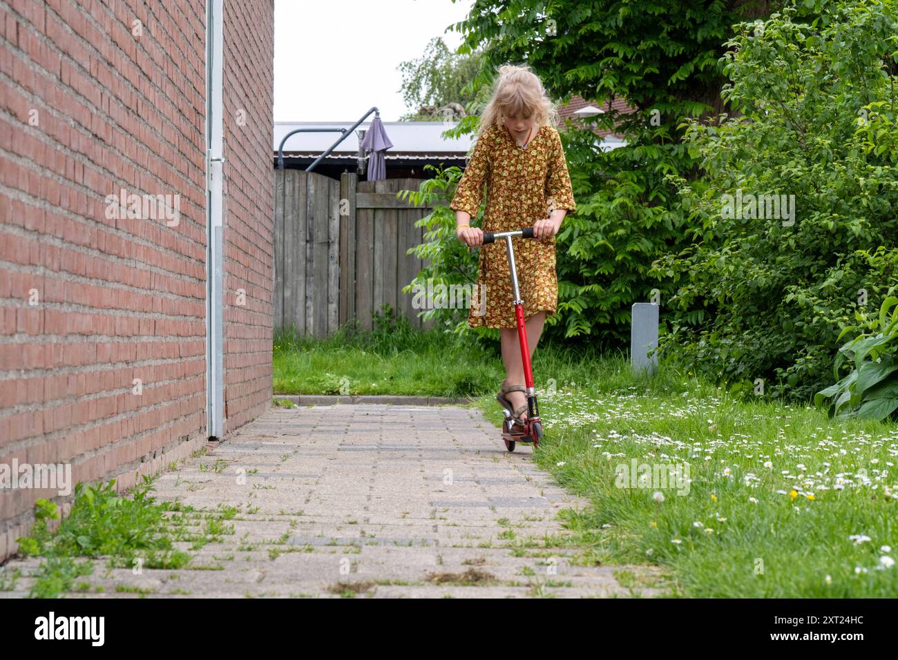 Young girl riding a scooter down a suburban pathway surrounded by green ...