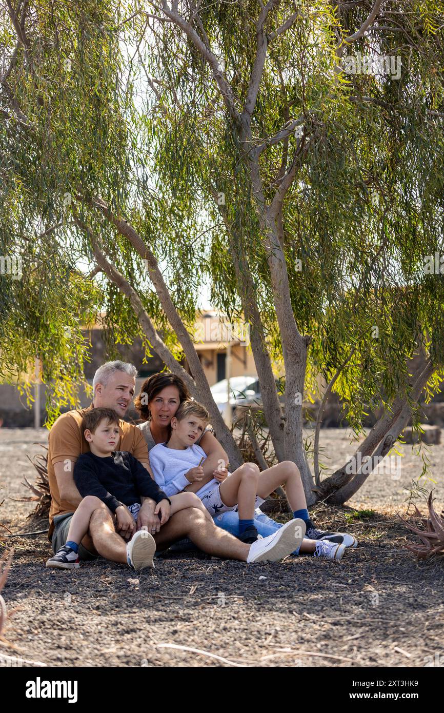 A family of four, mother, father, and two sons relaxes under the shade ...