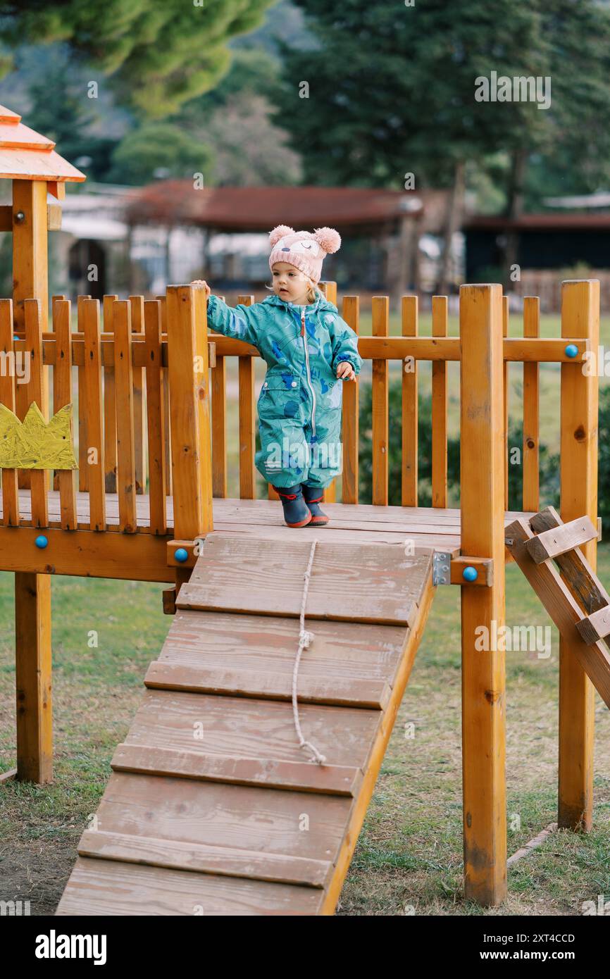 Little girl stands on top of a wooden climbing wall, holding on to the ...