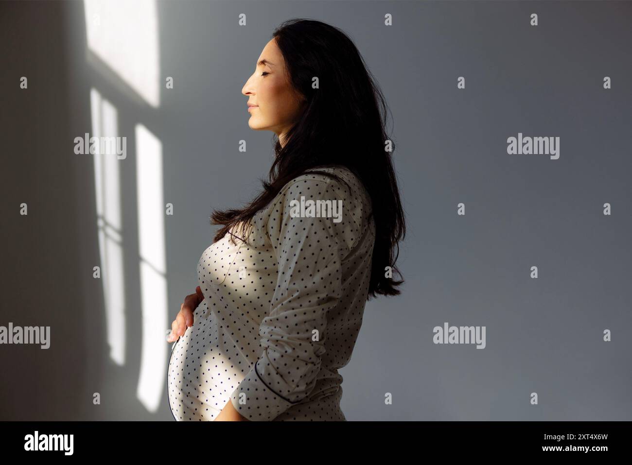 Pregnant woman stands in profile near window in dotted dress. Charming ...
