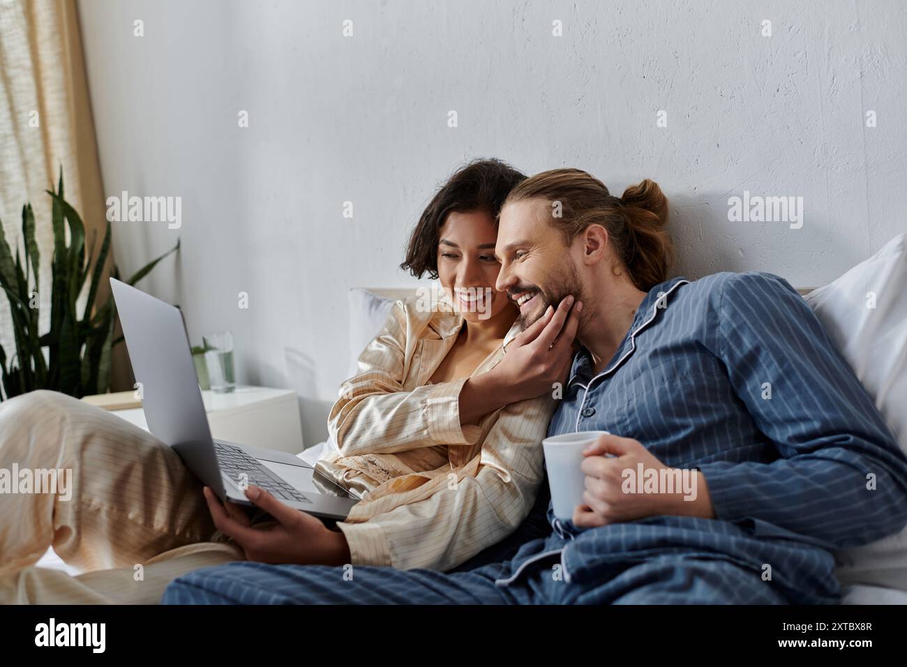 A couple cuddles in bed, enjoying a morning with coffee and a laptop ...