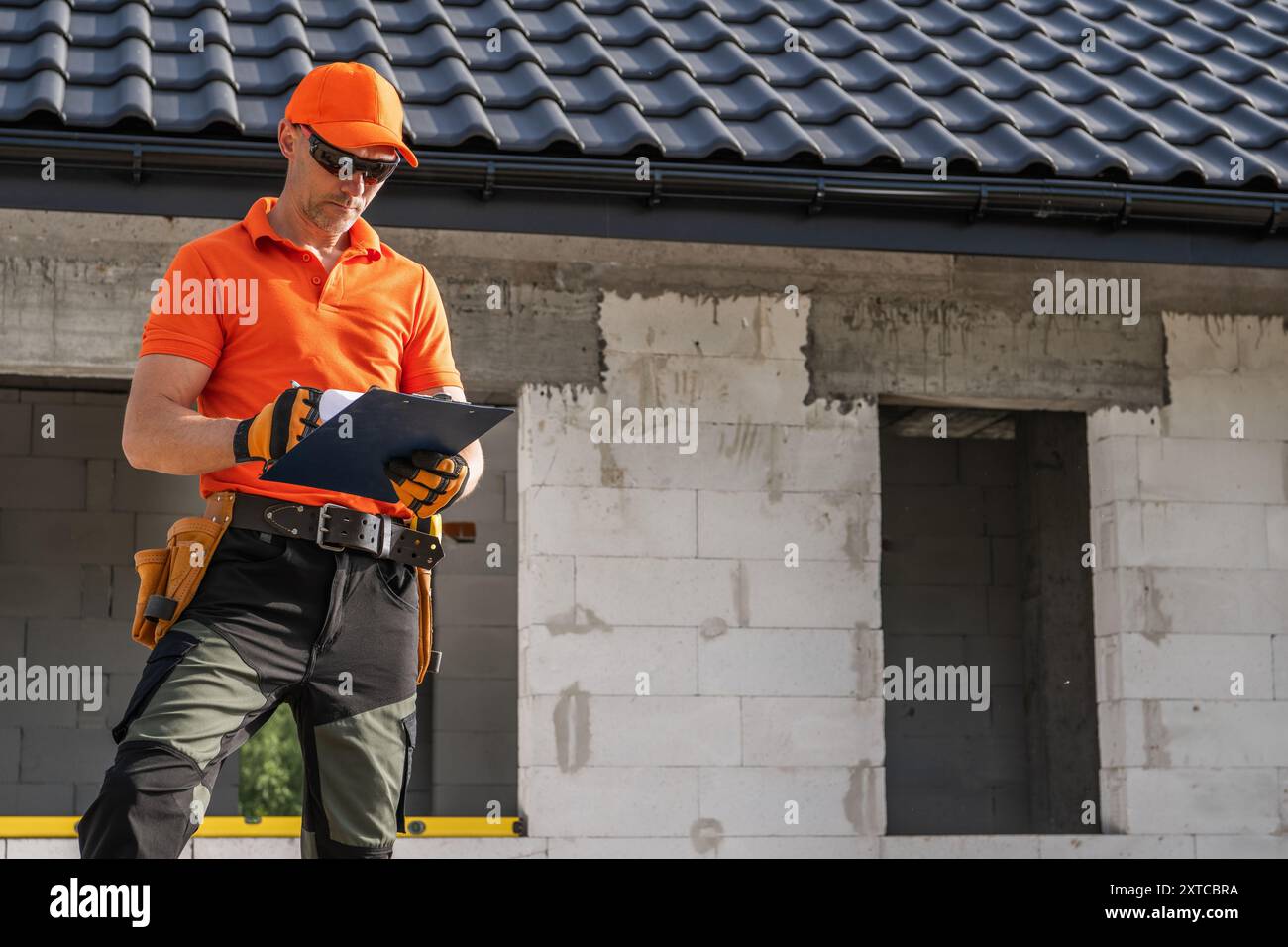 A construction worker in an orange outfit reviews a clipboard while overseeing the ongoing work ...