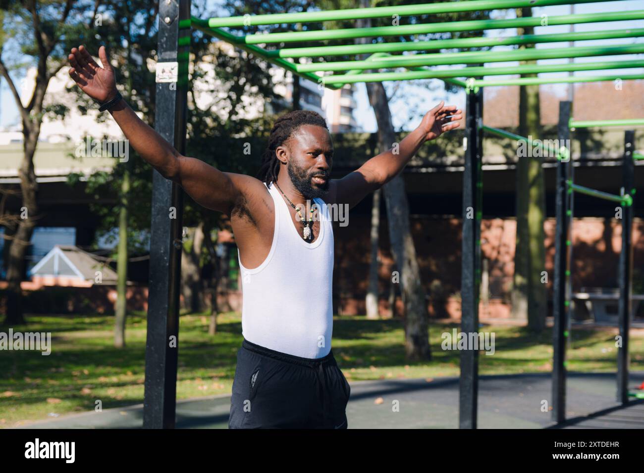 Black man with dreadlocks in sleeveless shirt stands outdoors with arms ...