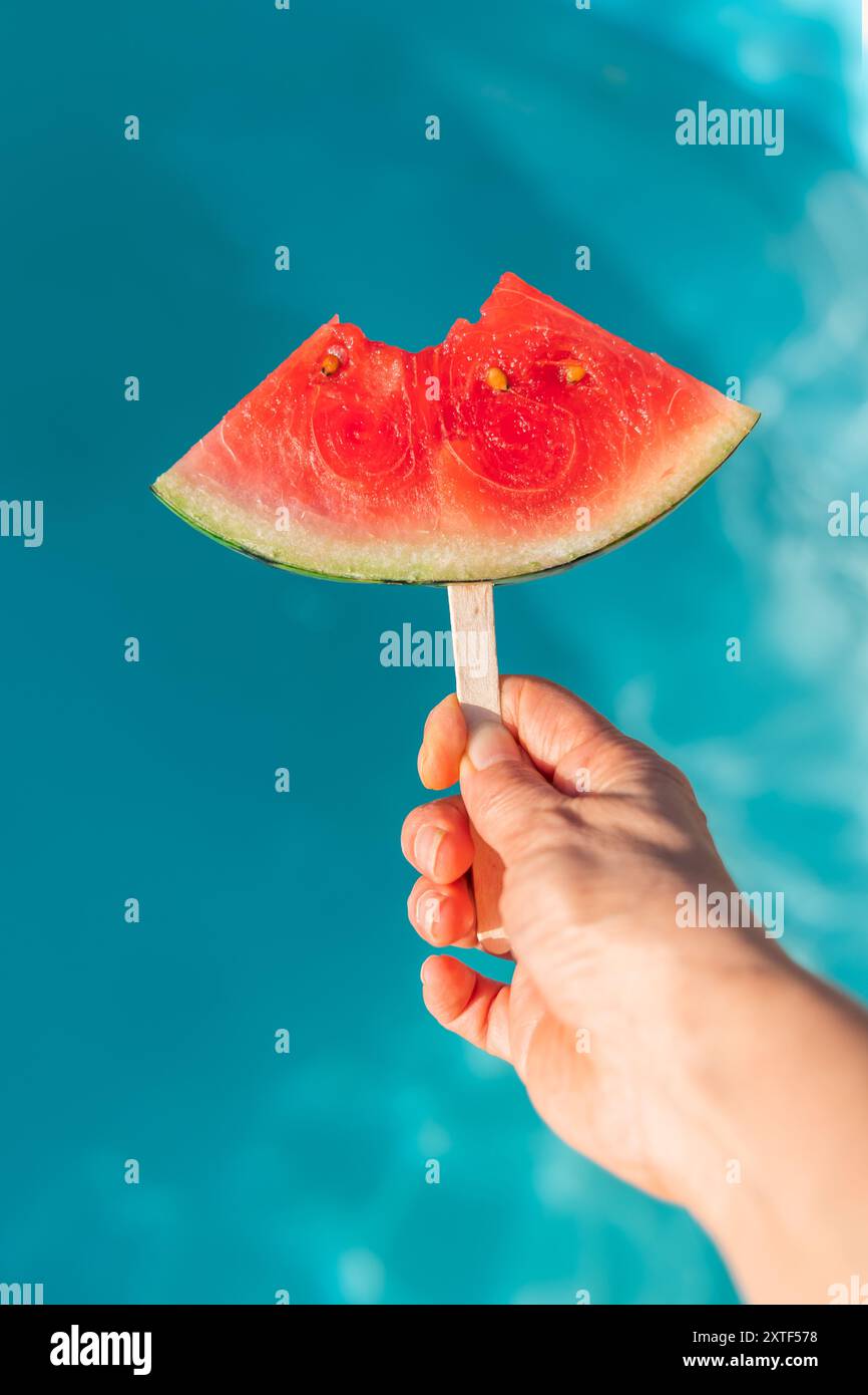 A hand holding a juicy watermelon slice on a stick above the calm blue ...