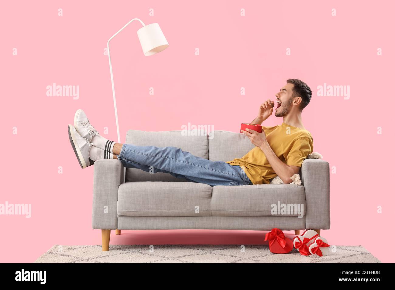Young man eating heart-shaped chocolate candies on sofa against pink ...