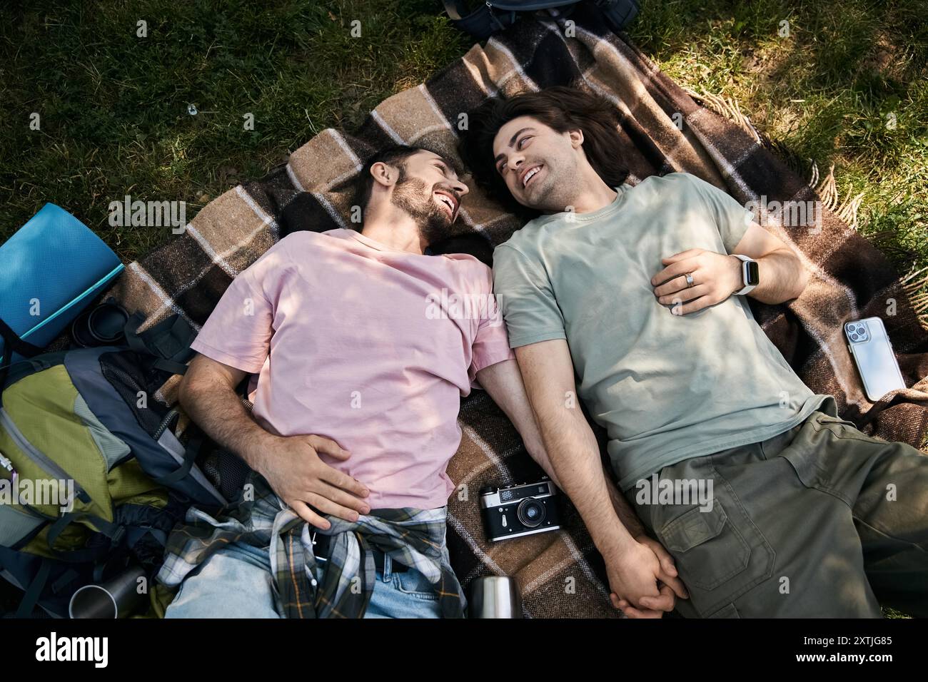 A young gay couple lies on a blanket in the wilderness, gazing lovingly ...