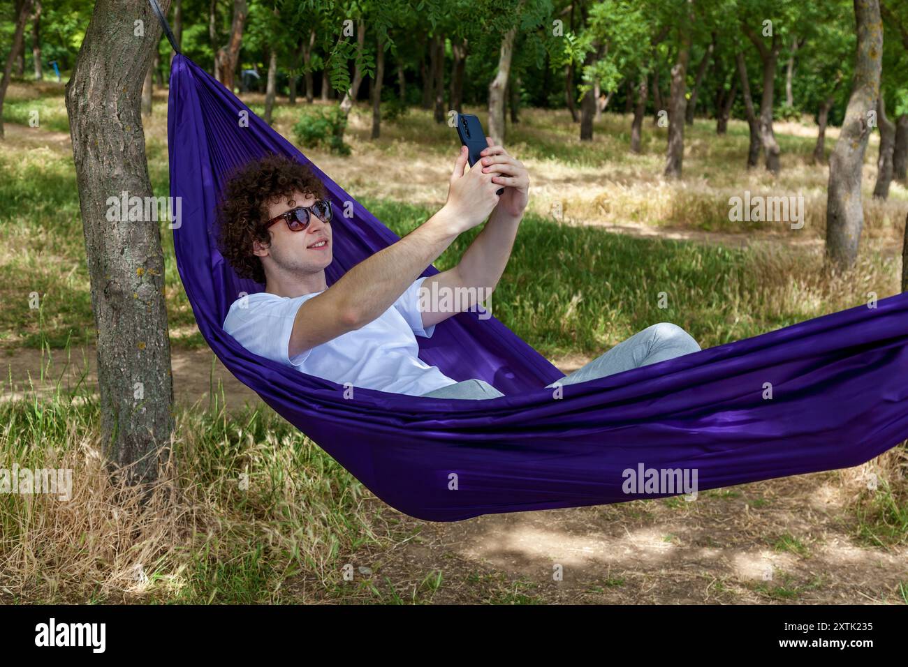 A young guy is lying in a purple hammock and taking a selfie on his ...