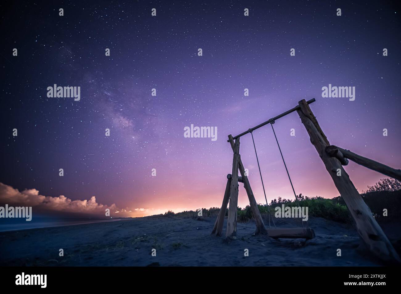 The Milky Way stretches across the night sky. A solitary wooden swing set stands on a sandy ...