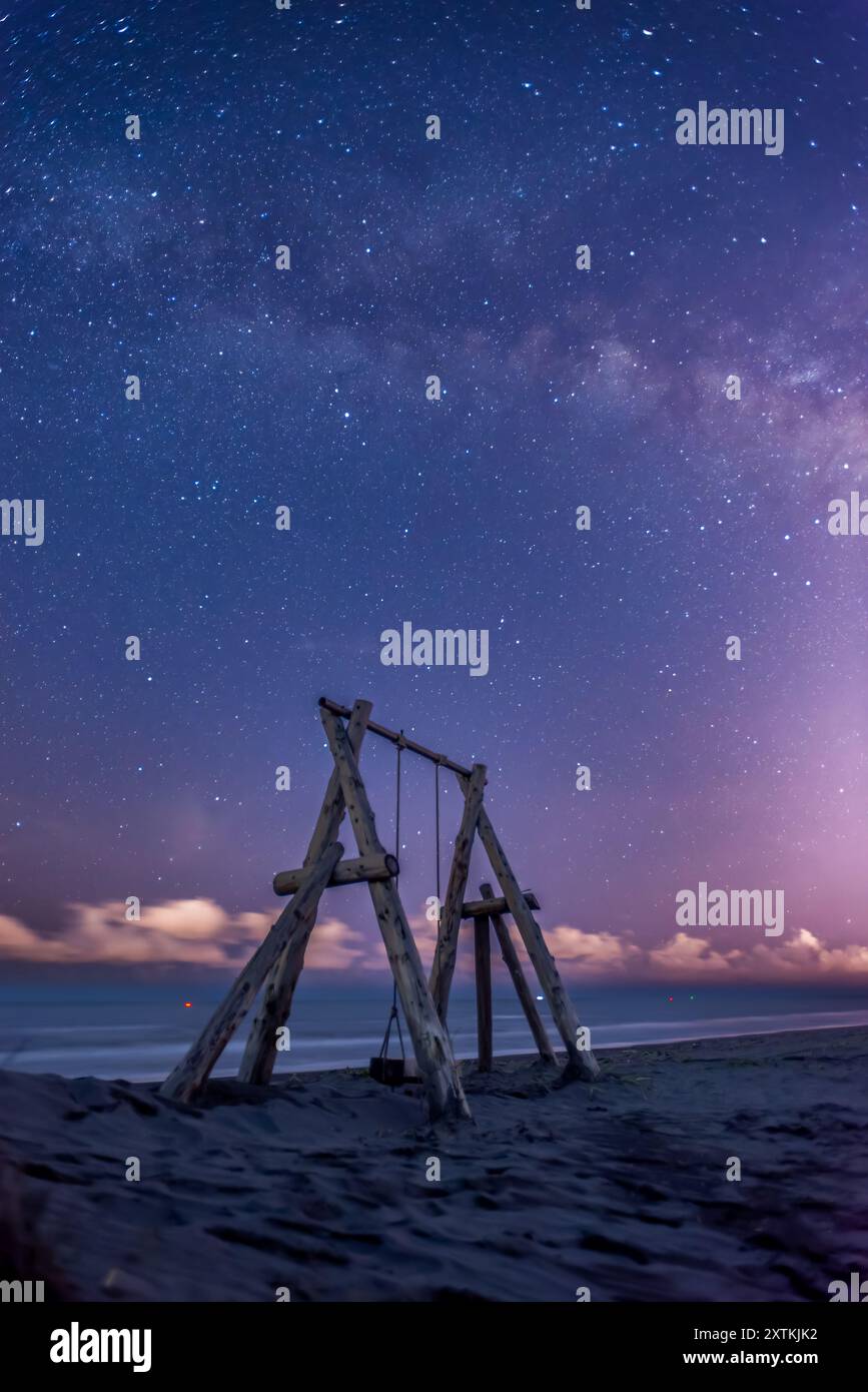 The Milky Way stretches across the night sky. A solitary wooden swing set stands on a sandy ...