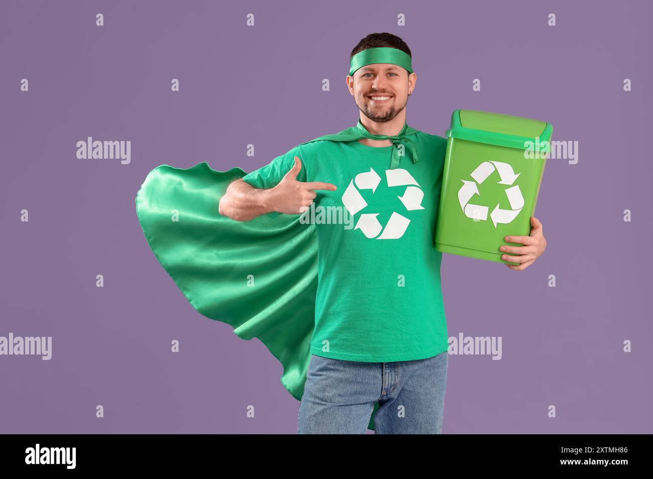 Young man dressed as eco superhero pointing at garbage bin on lilac ...