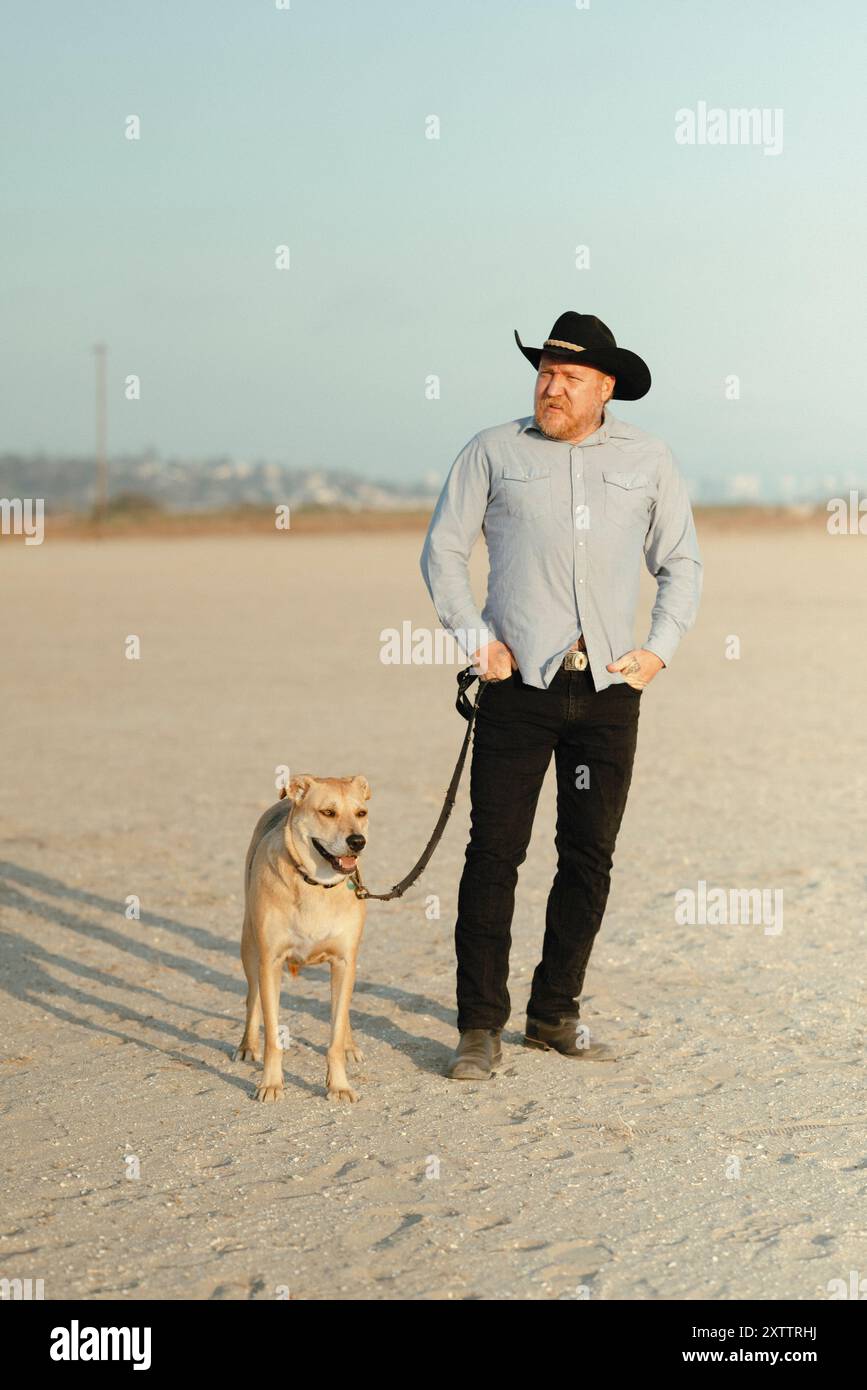 Man in a cowboy hat walking his dog on a sandy beach during sunset ...