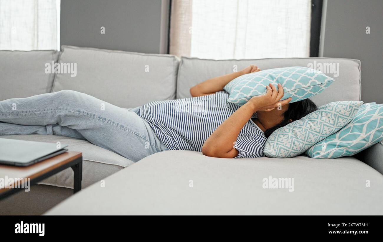 A stressed woman lying on a couch with a pillow over her head in a cozy ...