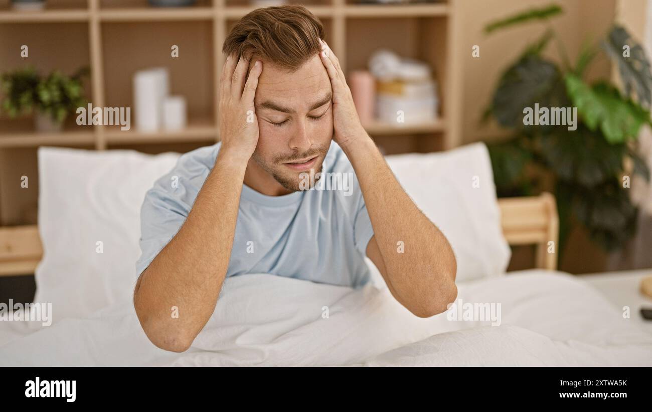 A troubled young man sitting on a bed in a neat bedroom, holding his ...