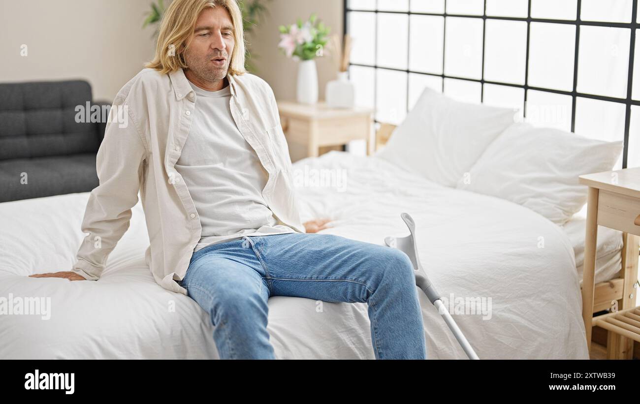 Injured young man with crutches sitting on the bed in a bright bedroom ...